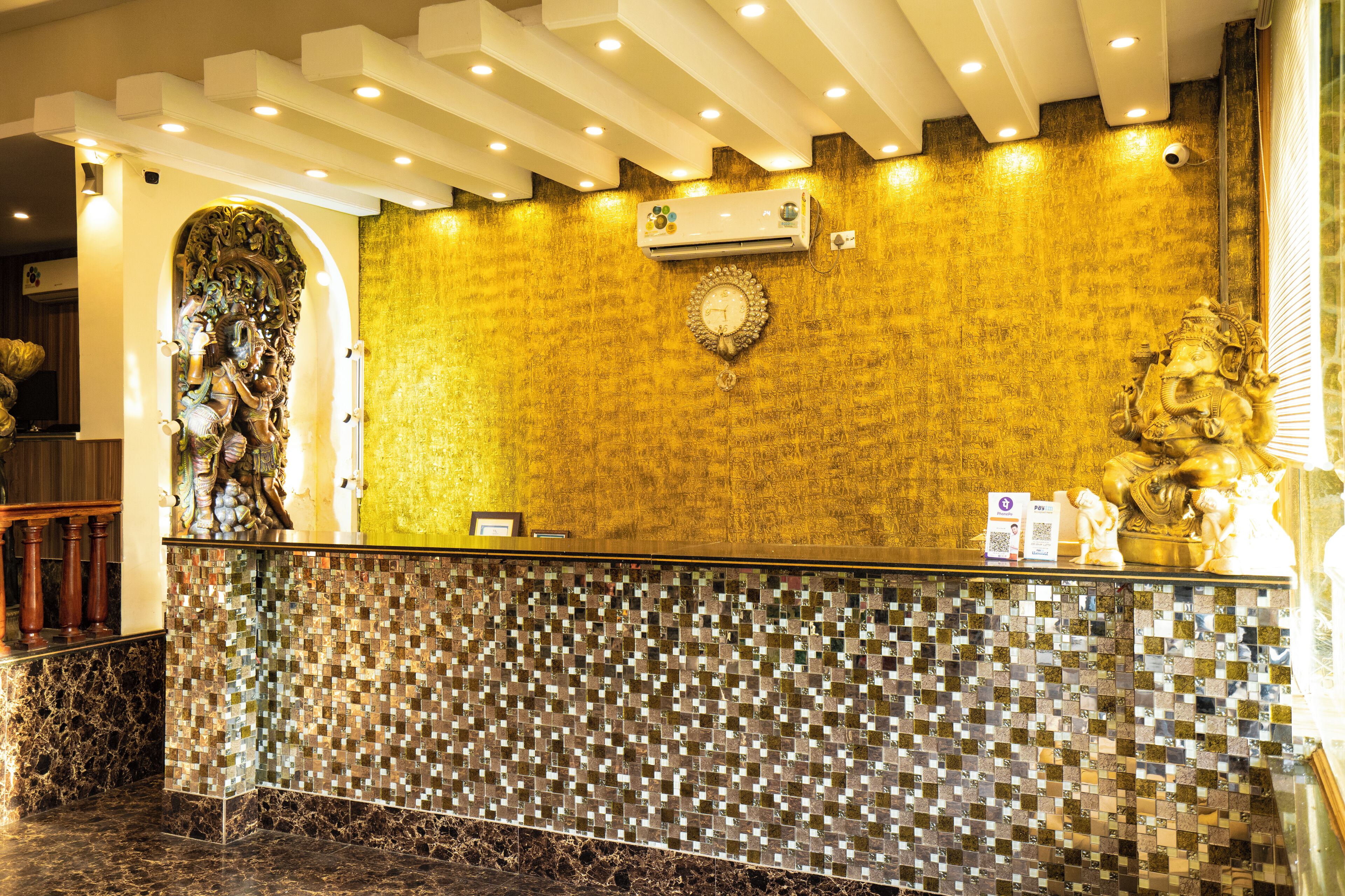 Uma área de recepção com balcão em mosaico, parede com textura dourada e uma estátua.