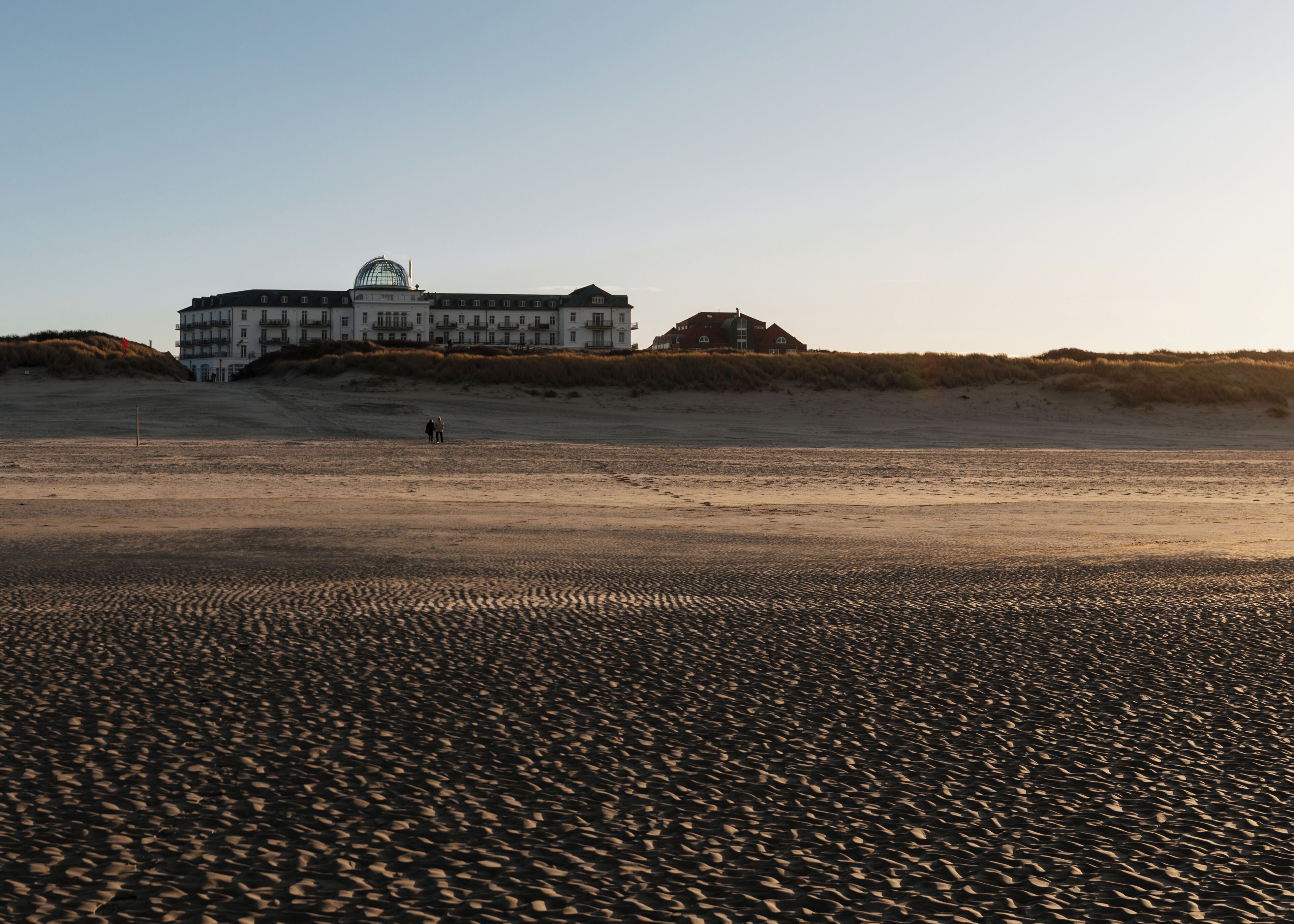 Um edifício grande com cúpula, localizado numa praia arenosa com dunas ao fundo.