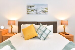 Uma cama bem arrumada com cabeceira, duas mesas de cabeceira com abajures, um quadro na parede e um pequeno relógio em uma das mesas.