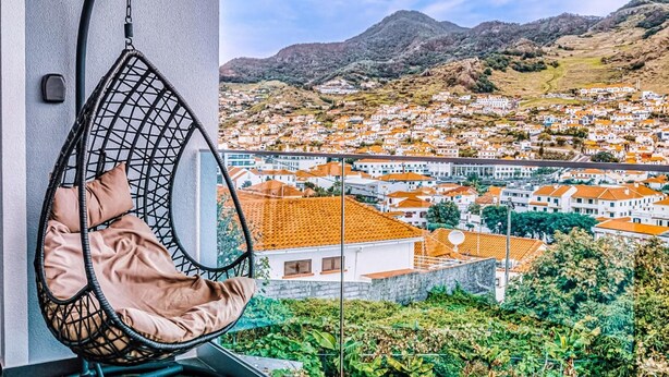 Uma varanda com uma cadeira suspensa, vista de uma cidade com edifícios de telhado vermelho e colinas verdes ao longe.