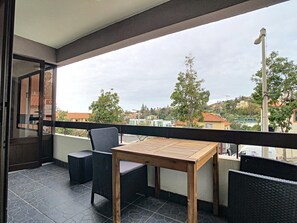 Varanda com mesa de madeira e duas cadeiras de vime, com vista para uma área residencial com casas e árvores.