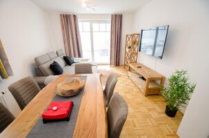 Uma sala de estar moderna com uma mesa de jantar de madeira, sofá cinza e uma TV fixada na parede.