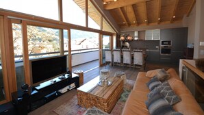Ampla sala de estar com uma TV grande, móveis de madeira e uma varanda com vista para as montanhas.