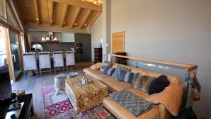 Uma sala de estar ampla com um sofá grande, uma mesa de centro de madeira e uma mesa de jantar com cadeiras. Uma cozinha é visível ao fundo.