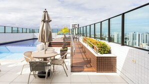 Área de piscina no terraço com vista para o horizonte da cidade, assentos externos e um pequeno jardim.