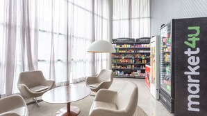 Um saguão de hotel com área de estar, uma mesa, um abajur, um refrigerador e uma máquina de vendas.