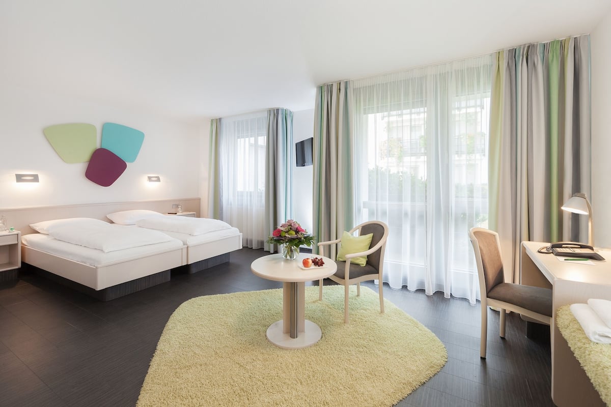 Quarto de hotel com cama, escrivaninha, cadeira, televisão e uma pequena mesa com um vaso de flores.