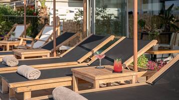 Área de piscina com espreguiçadeiras e mesas de madeira, algumas com toalhas e bebidas.