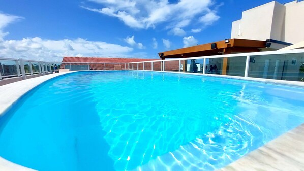 Uma piscina azul cristalina com um deck branco, rodeada por um guarda-corpo de vidro e um edifício com telhado vermelho ao fundo.