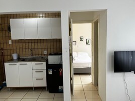 Uma cozinha compacta com armários brancos, uma pia e uma geladeira pequena. Uma televisão está instalada na parede do quarto vizinho.