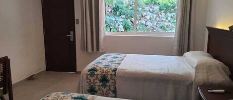 Quarto de hotel com duas camas, uma janela com cortinas, uma porta e uma pequena mesa com um controle remoto.