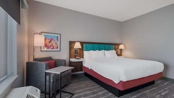 Quarto de hotel com cama, escrivaninha, cadeira, luminária e um quadro na parede.