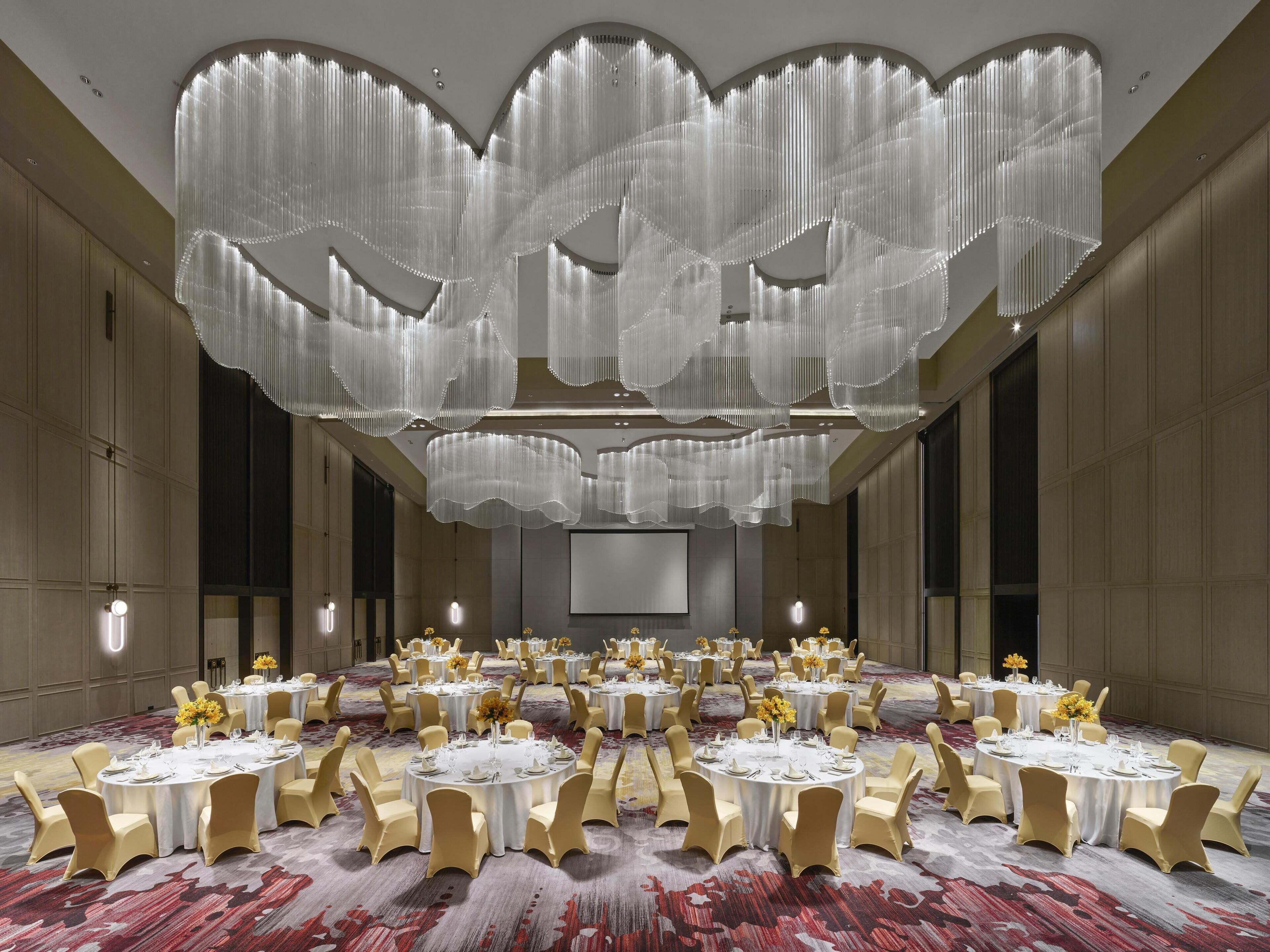 Uma grande sala de conferências com várias mesas redondas, cada uma com toalhas brancas e cadeiras douradas, e um teto com um design diferenciado que suaviza a luz.