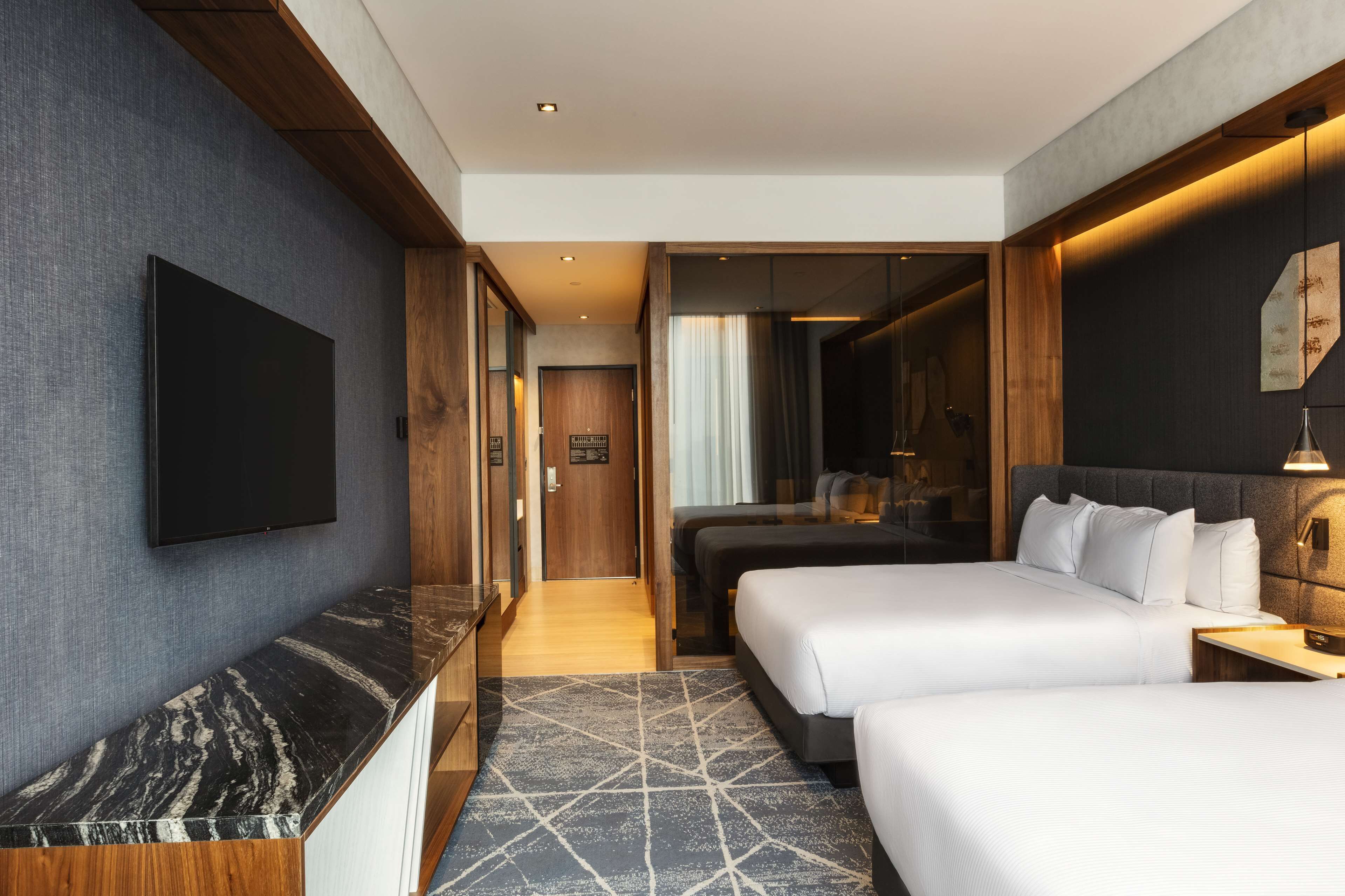 Quarto de hotel moderno com TV de tela plana na parede, duas camas com roupa de cama branca, cabeceira de madeira, bancada de mármore e tapete estampado.