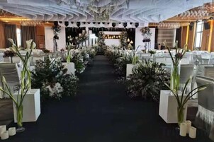 Um longo corredor com mesas e cadeiras, decorado com folhagem e flores brancas, que leva a uma área de bar.