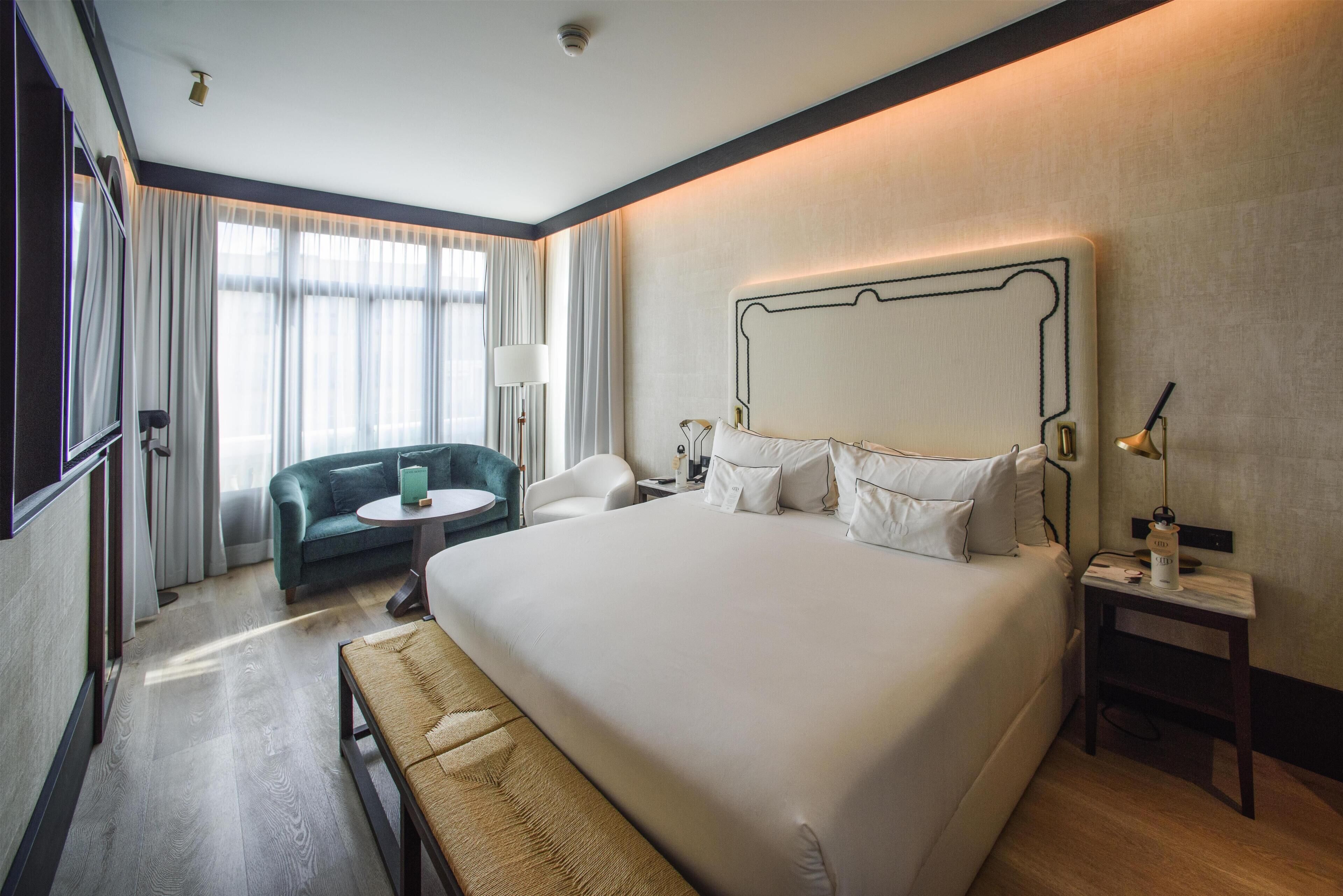 Quarto de hotel com uma cama grande, um sofá, uma mesinha redonda, um banquinho e uma mesa de cabeceira com abajur.