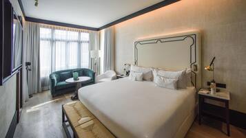 Quarto de hotel com uma cama grande, um sofá, uma mesinha redonda, um banquinho e uma mesa de cabeceira com abajur.