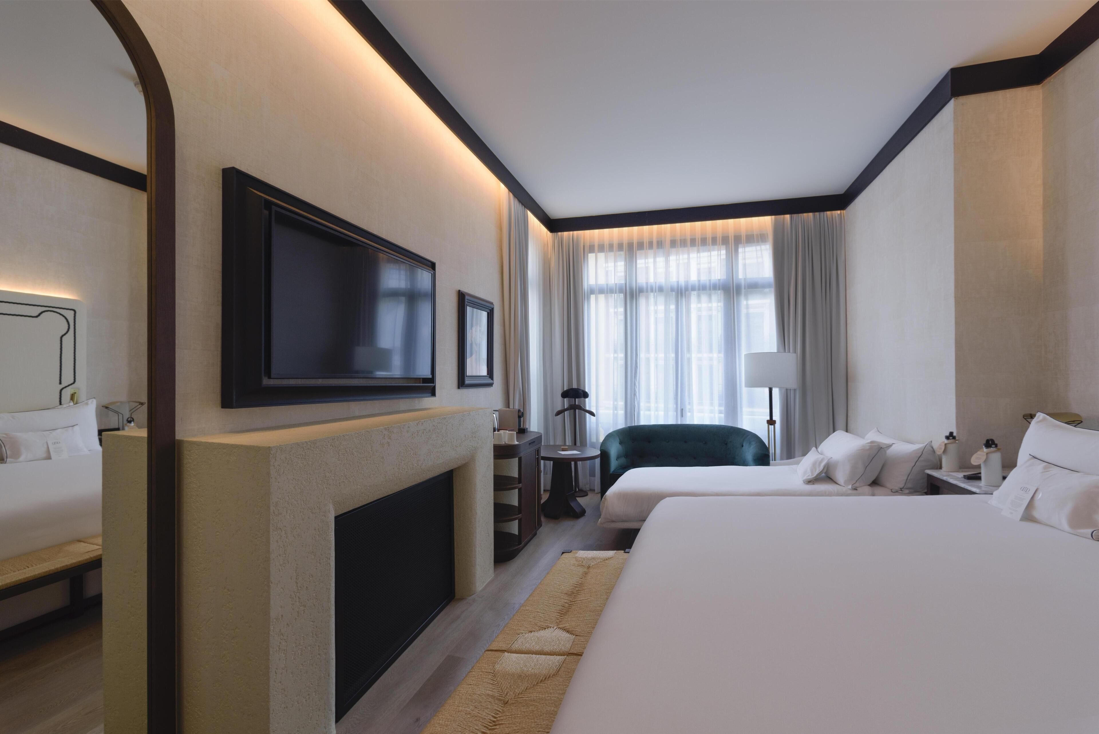 Quarto de hotel com televisão, duas camas, lareira, escrivaninha e janela com cortinas.
