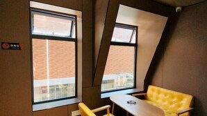 Quarto com sofá amarelo, uma mesa pequena e duas janelas com vista para um edifício de tijolos.