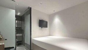 Quarto de hotel moderno com cama, televisão fixada na parede e porta de vidro para um banheiro.