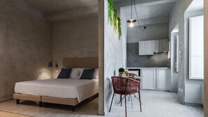 Quarto com cama, mesa de cabeceira e uma cadeira. Cozinha visível com armários e bancada.