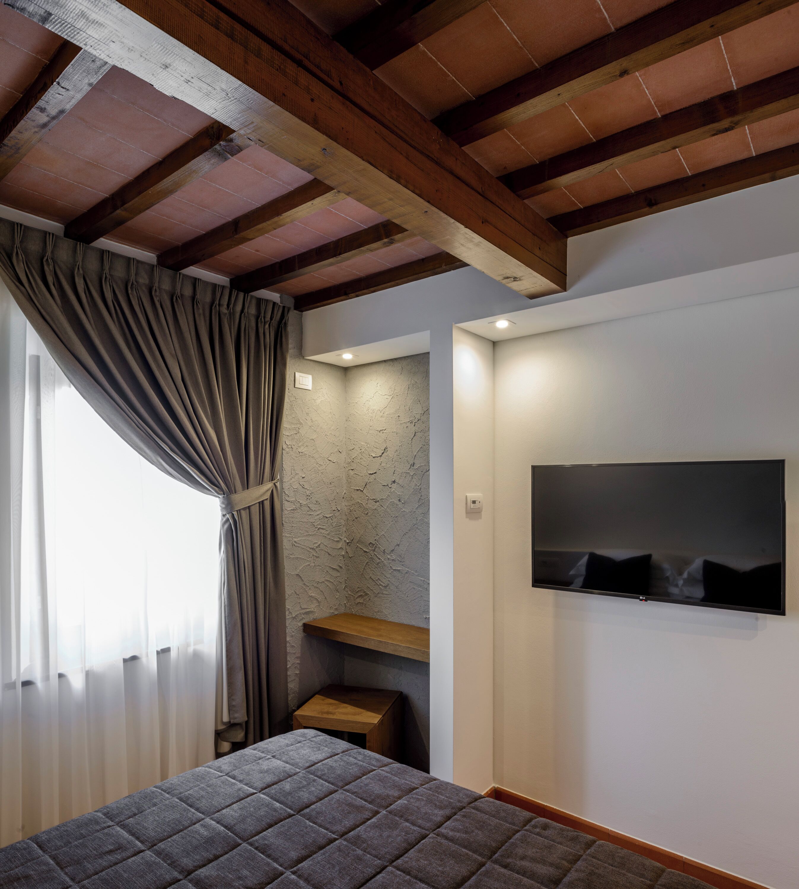 Quarto com cama, televisão presa na parede, um banco de madeira e janela com cortinas.