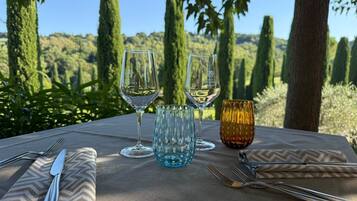 Uma mesa posta ao ar livre, com taças de vinho e talheres, rodeada por vegetação e árvores.