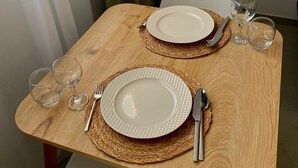 Mesa de jantar posta com pratos, talheres e copos, com uma cadeira e um vaso com plumas de lagosta ao fundo.