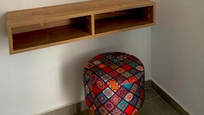 Um espelho redondo com moldura preta, uma prateleira de madeira abaixo e um banquinho colorido à direita.