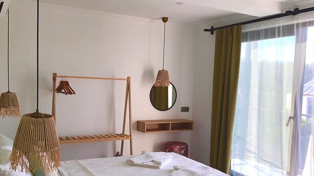 Quarto de hotel com uma cama, mesa de cabeceira de madeira, luminária pendente, espelho e janela com cortinas.