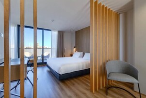 Quarto de hotel moderno com uma cama grande, uma mesa e uma cadeira. Vê-se a cidade através de amplas janelas.