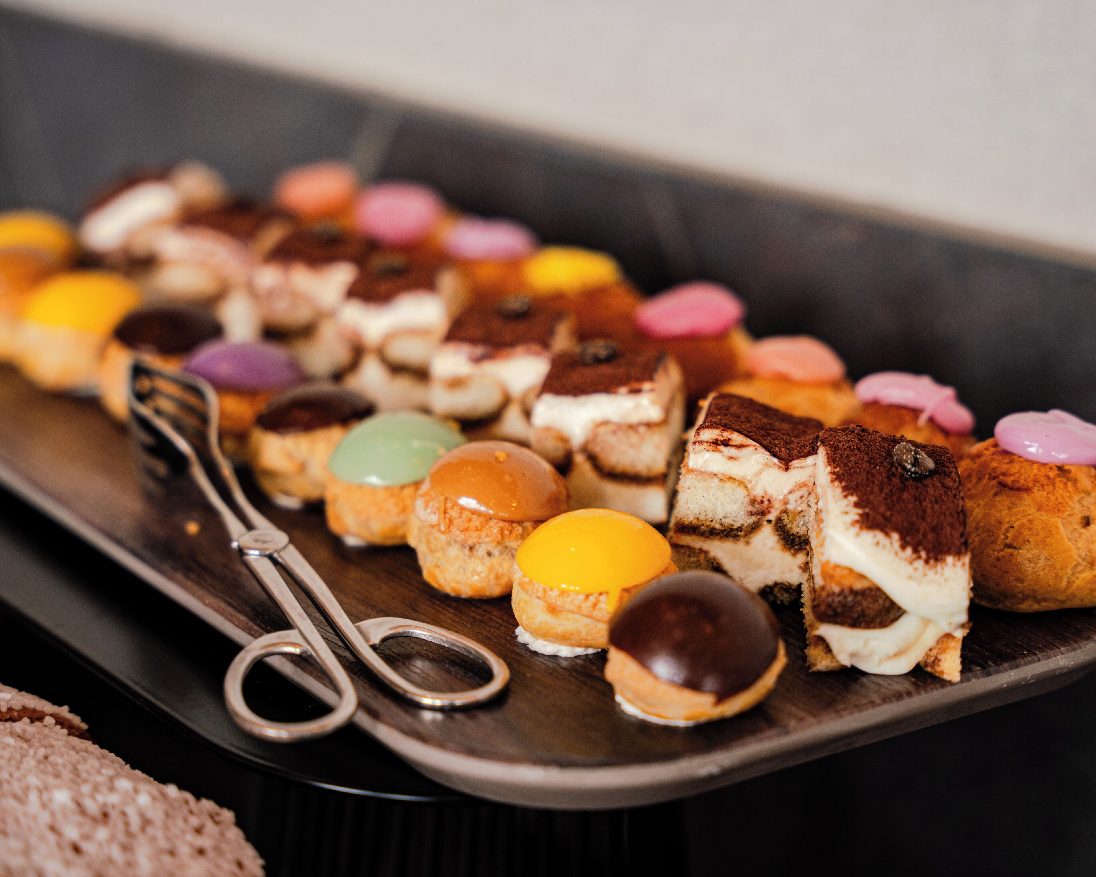 Uma bandeja com diversos doces e sobremesas, incluindo bolos e biscoitos, com um jogo de pinças ao lado.