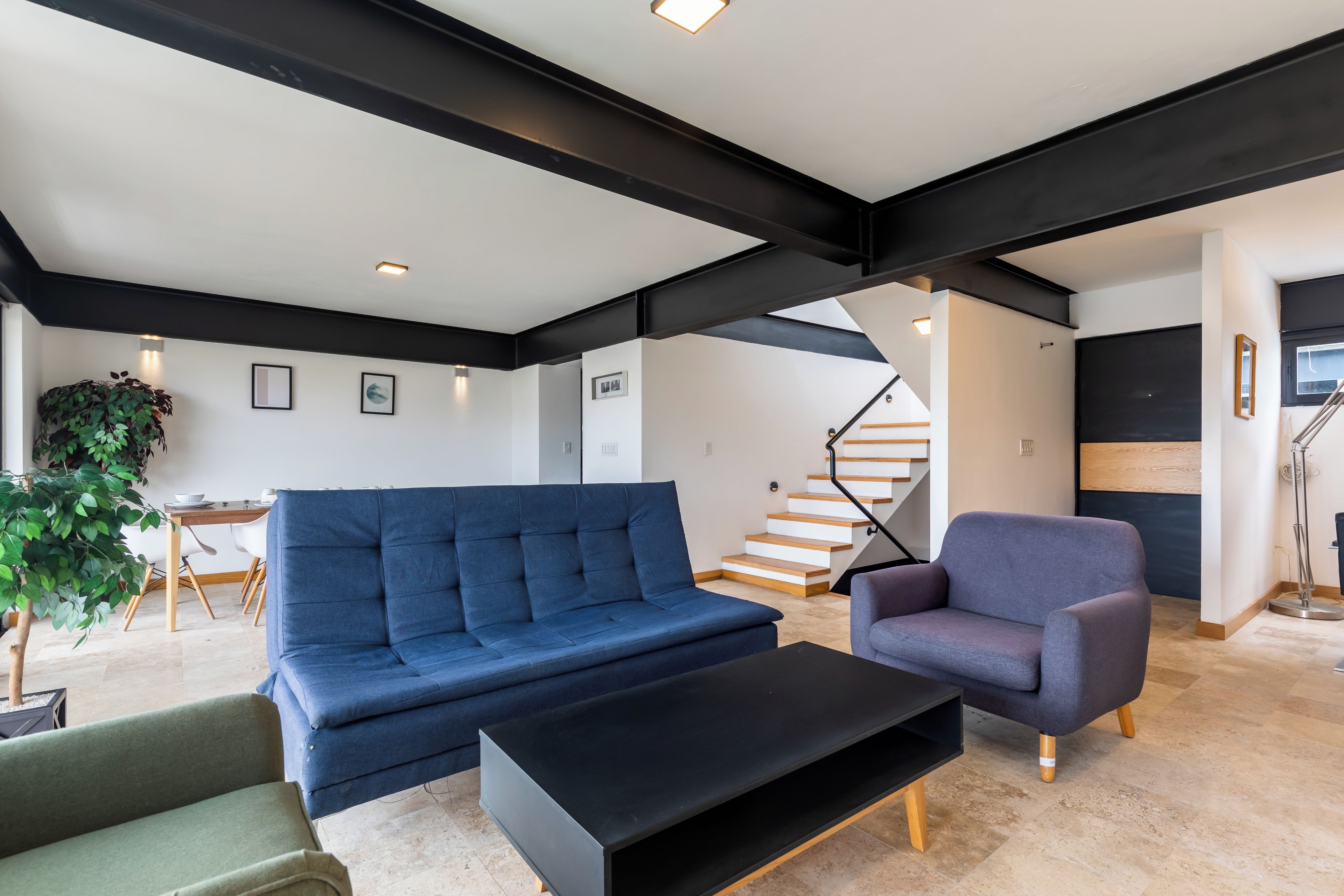 Uma sala de estar moderna com um sofá azul, uma poltrona cinza e uma mesa de centro preta. Uma escada ao fundo leva a um andar superior.
