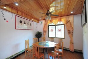 Uma área de refeições com uma mesa redonda, cadeiras de madeira, ventilador de teto e uma janela com cortinas.