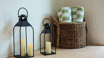 Dois castiçais estilo lanterna em preto com velas, uma cesta de palha com almofadas quadriculadas verdes e brancas, e uma parede lisa.