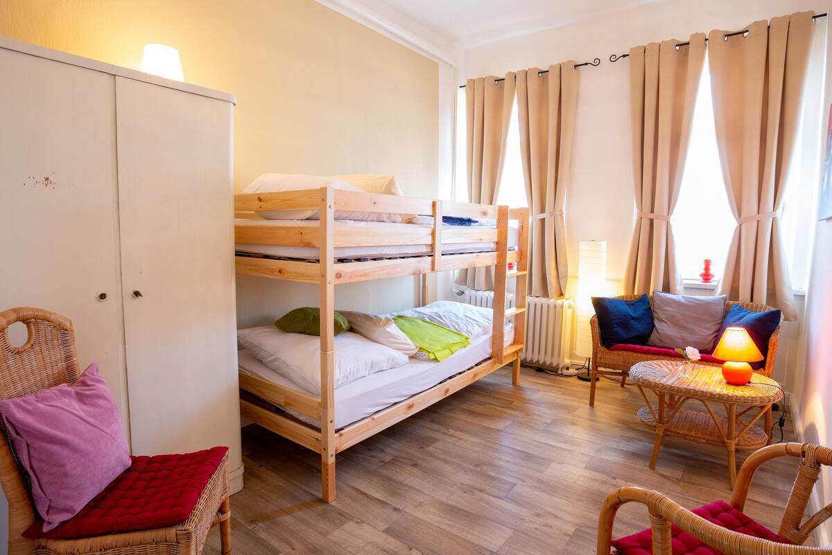 Uma cama beliche com estrutura de madeira, uma cadeira de vime com almofada vermelha, uma mesa de vime com abajur e um radiador.
