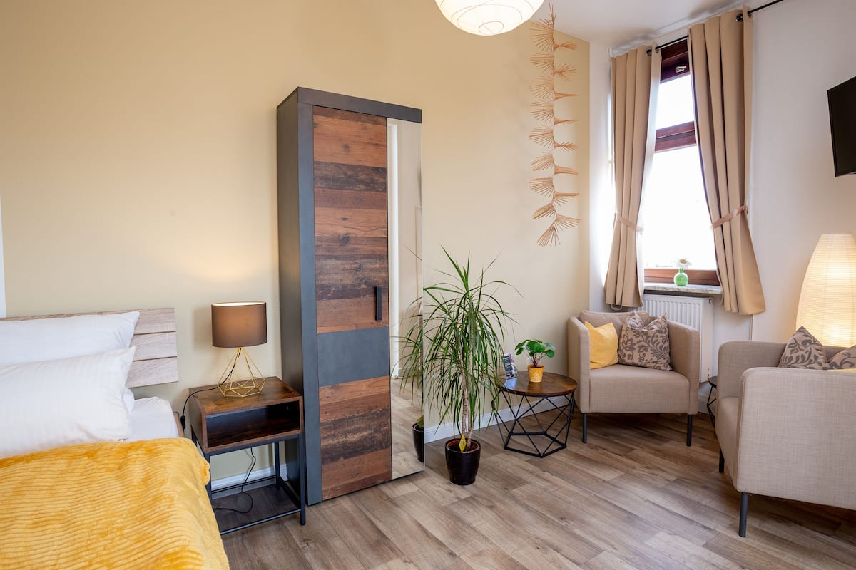 Quarto de hotel moderno com cama, sofá, mesinha, planta em vaso e janela com cortinas.
