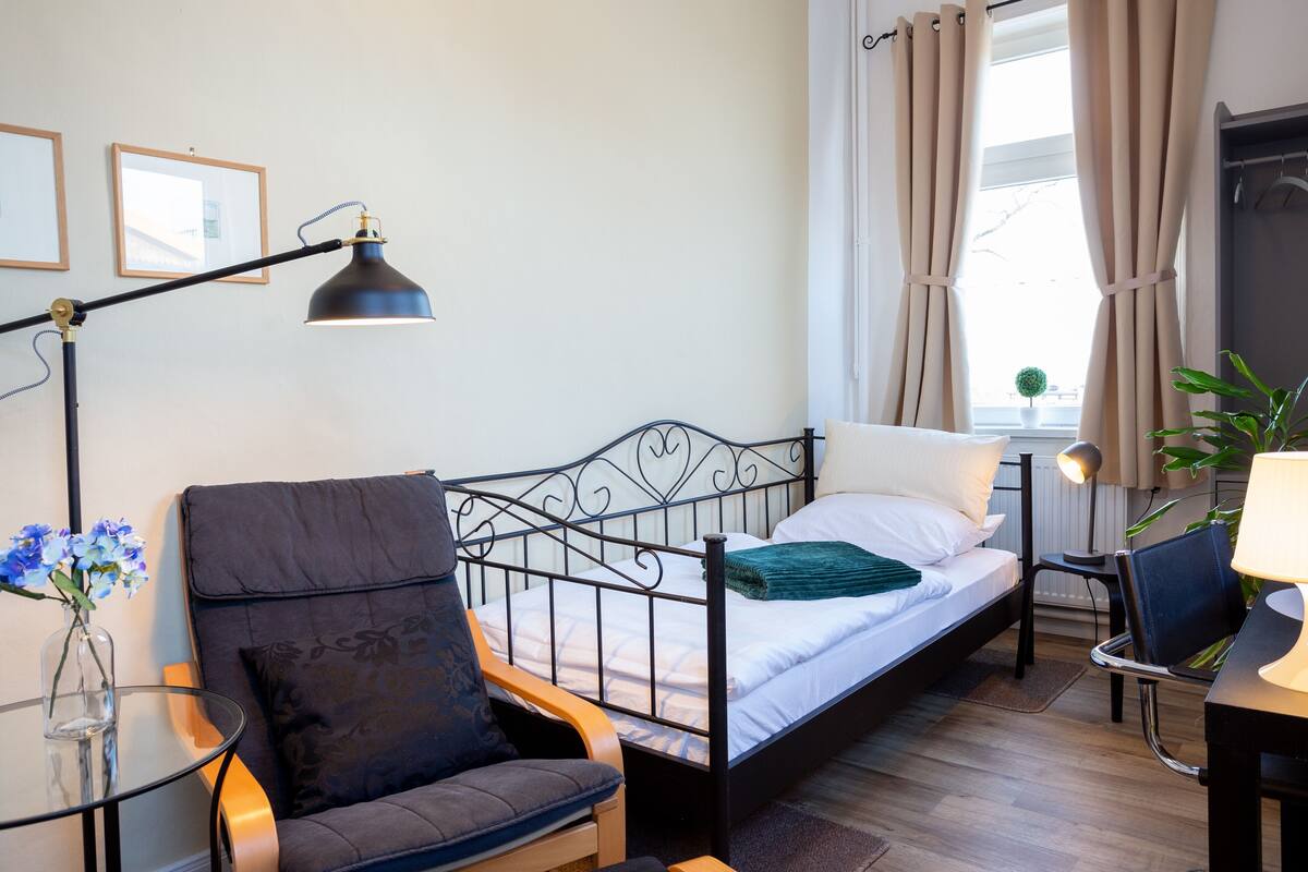 Quarto com uma cama de solteiro, uma cadeira, uma escrivaninha, uma luminária e uma janela com cortinas.