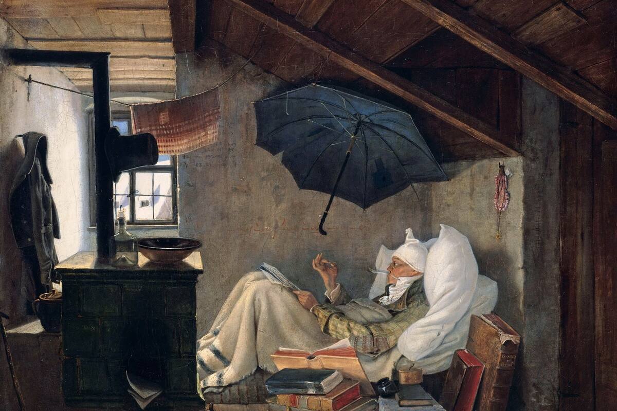 Um quarto com teto de madeira, uma janela, uma mesa com livros e uma pessoa lendo sob um guarda-chuva.