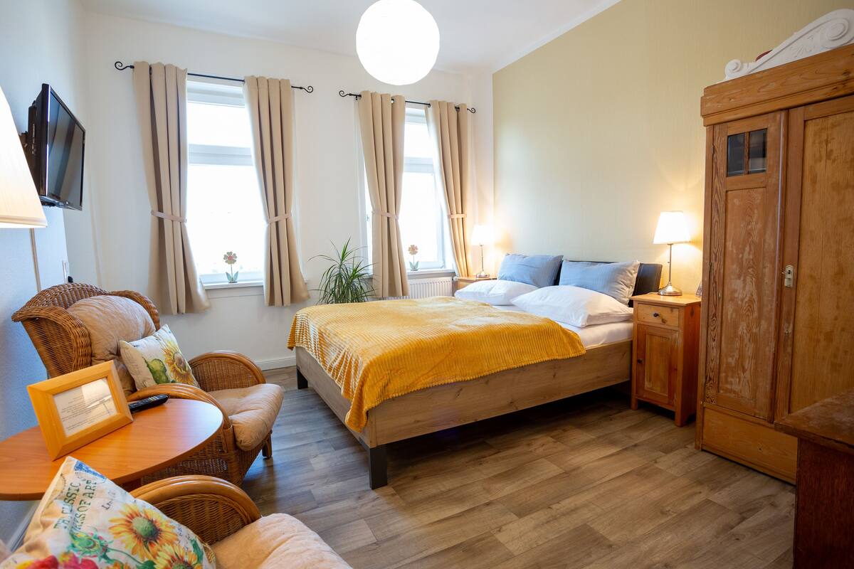 Um quarto com cama, uma cadeira, mesa de cabeceira, guarda-roupa e televisão na parede.