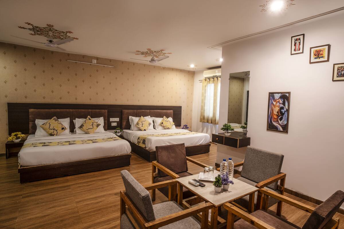 Quarto de hotel com duas camas, uma mesa de jantar com cadeiras, uma mesinha com um vaso e comando remoto, e obras de arte nas paredes.