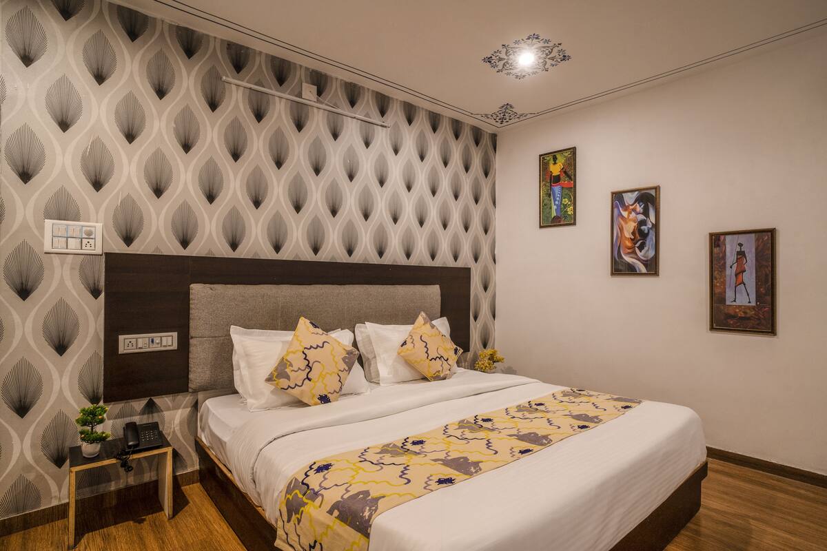 Quarto de hotel com papel de parede estampado, cama com colcha estampada, mesa de cabeceira com telefone e uma pequena planta, e obras de arte nas paredes.