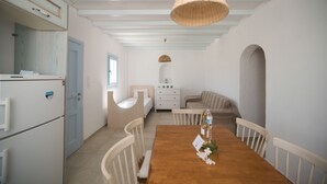 Quarto compacto com mesa de jantar de madeira, cadeiras brancas, pequeno espaço de cozinha, cama e luminária pendente.