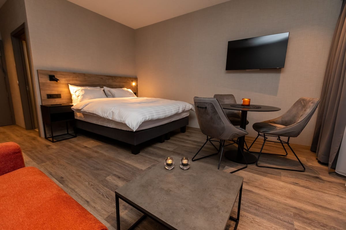 Quarto de hotel moderno com cama, escrivaninha, duas cadeiras, televisão e pequena mesa com velas.