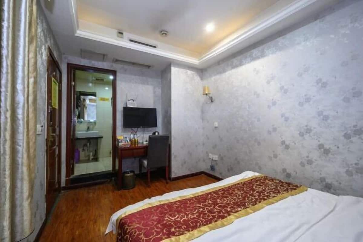 Quarto de hotel com cama, escrivaninha, cadeira, televisão e banheiro visível por uma porta aberta.