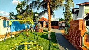 Um parquinho com balanços e um carrossel amarelo, cercado por palmeiras e casas com telhados vermelhos.