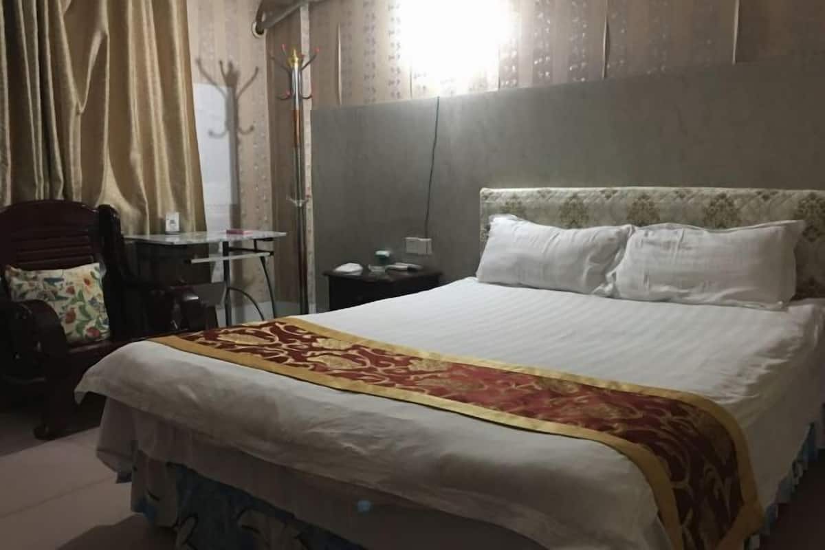 Uma cama bem arrumada com colcha estampada, uma mesa de cabeceira com abajur e uma cadeira com almofada florida.