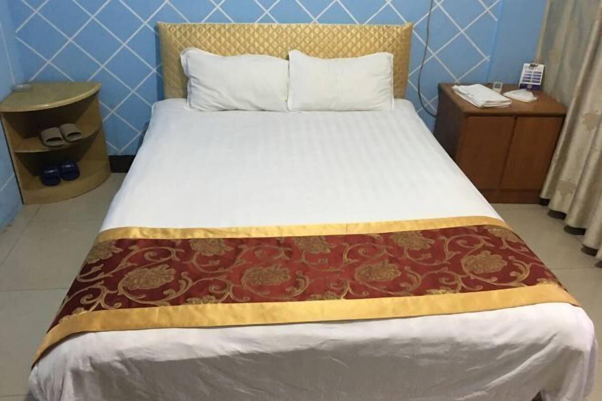 Cama bem arrumada com colcha estampada e almofadas, mesa de cabeceira com telefone e bloco de notas, e um pequeno estante com sapatos.