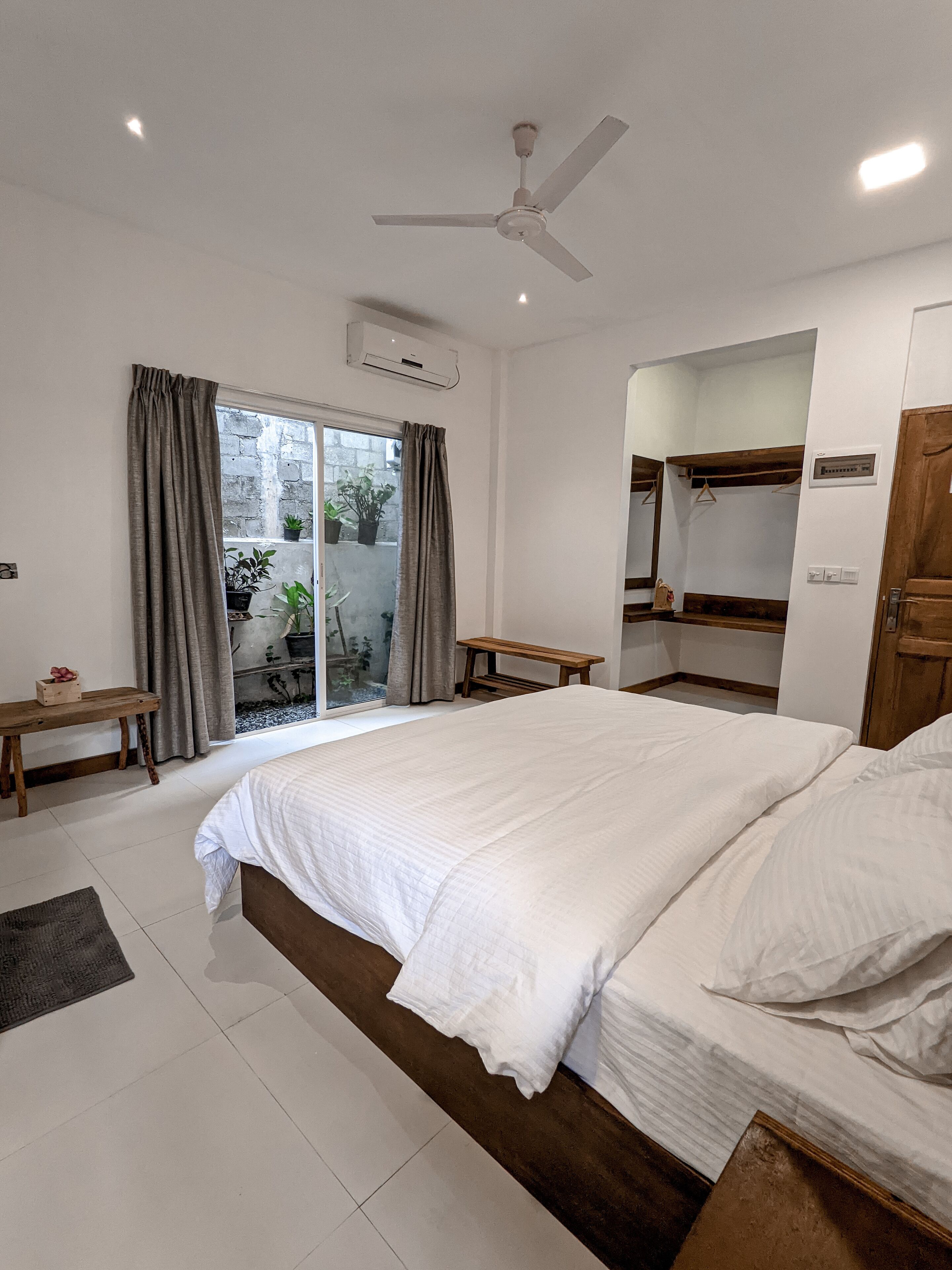 Quarto com cama grande, ventoinha no teto, banco de madeira, mesinha, espelho e porta de vidro deslizante para área externa com plantas.