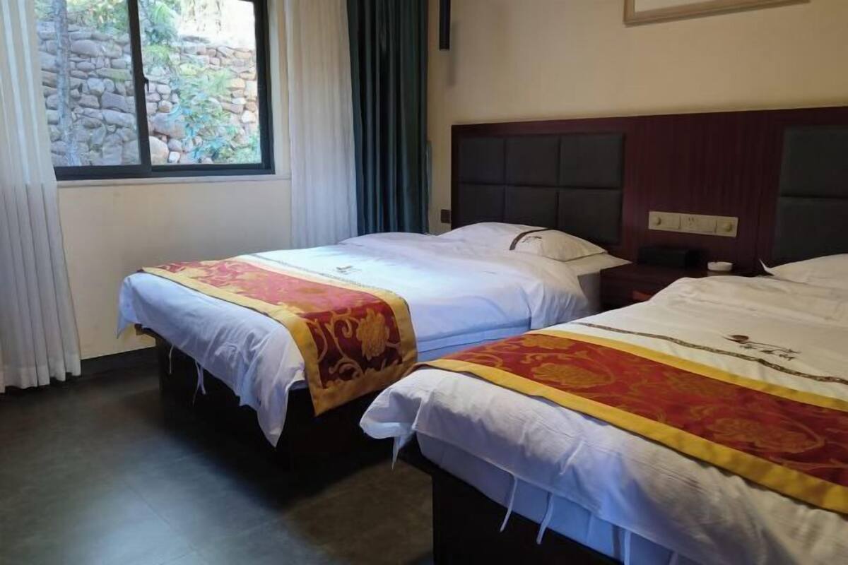 Quarto de hotel com duas camas, uma janela com cortinas, uma parede de pedra do lado de fora e uma mesa de cabeceira com abajur.